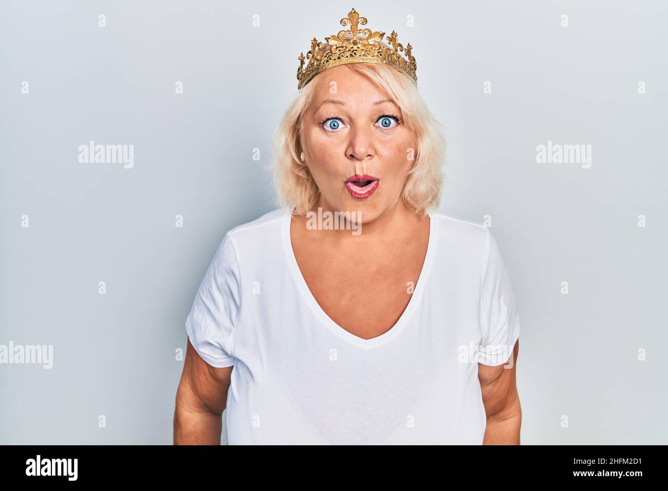 Middle age blonde woman wearing queen crown scared and amazed with open ...