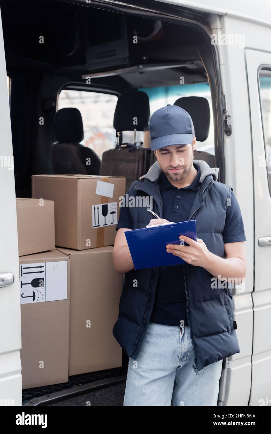 Young courier in uniform writing on clipboard near boxes with signs in ...