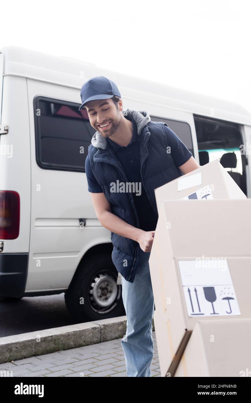 Smiling courier looking at carton boxes with signs near auto outdoors ...