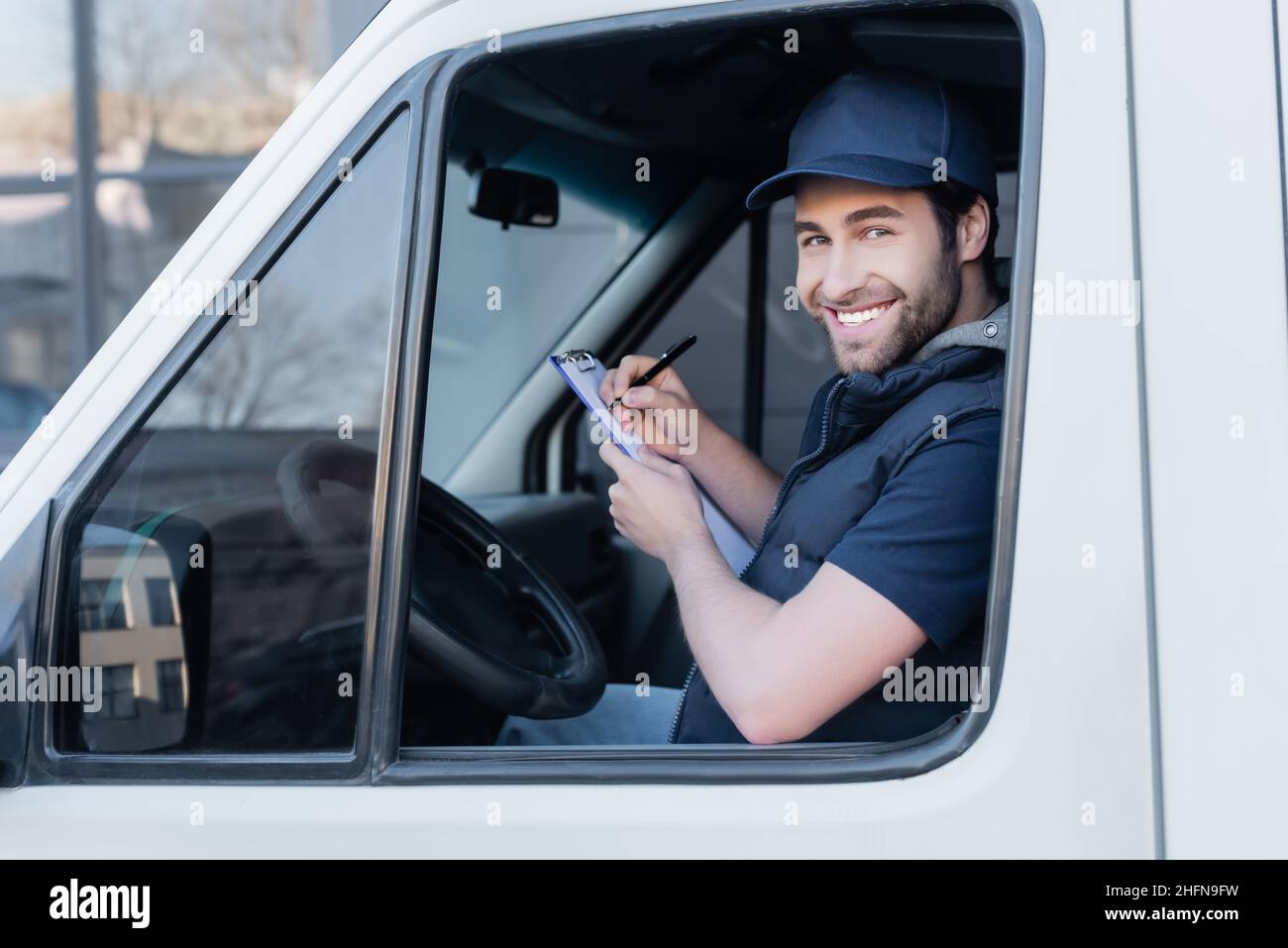 Young courier smiling at camera while writing on clipboard in car Stock ...