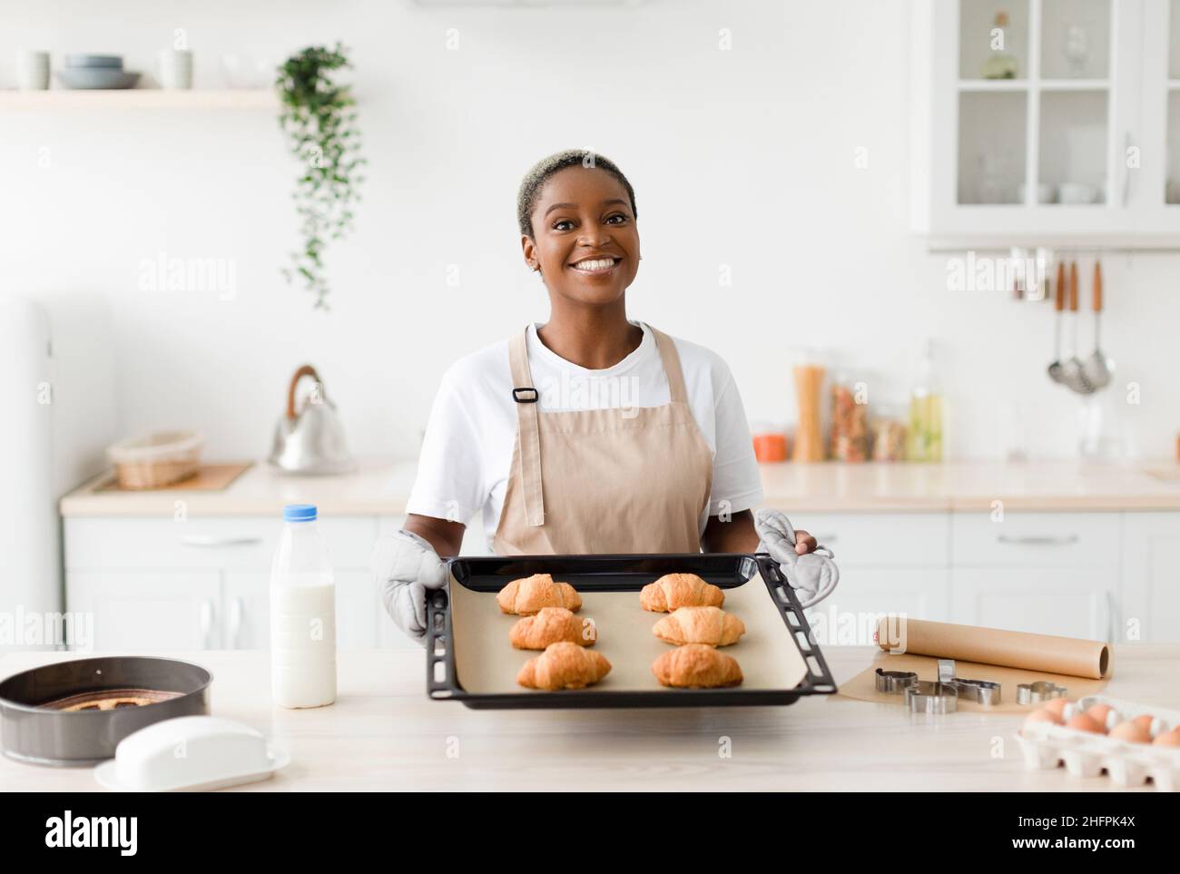 Satisfied young cute black woman in apron shows ready croissants in ...