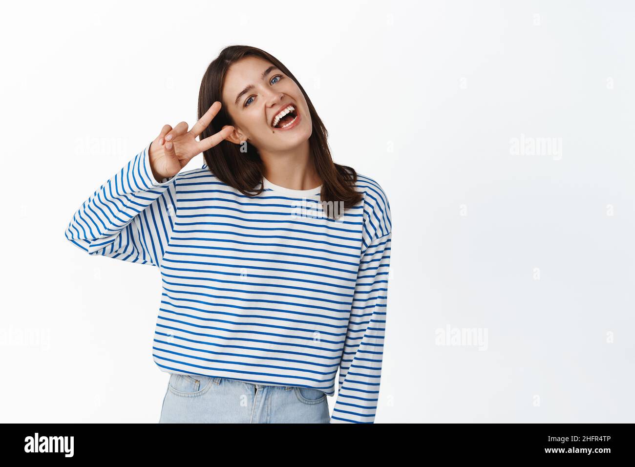 Happy And Positive Brunette Girl Shows Peace Sign And Smiles Laughing Carefree Standing In