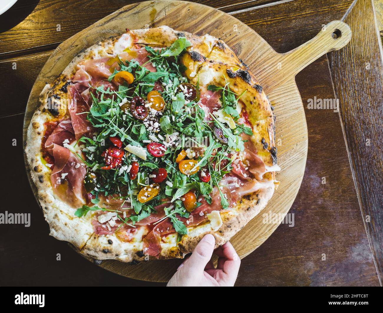 Man's hand takes slice of fresh pizza with ham, tomatoes and arugula ...