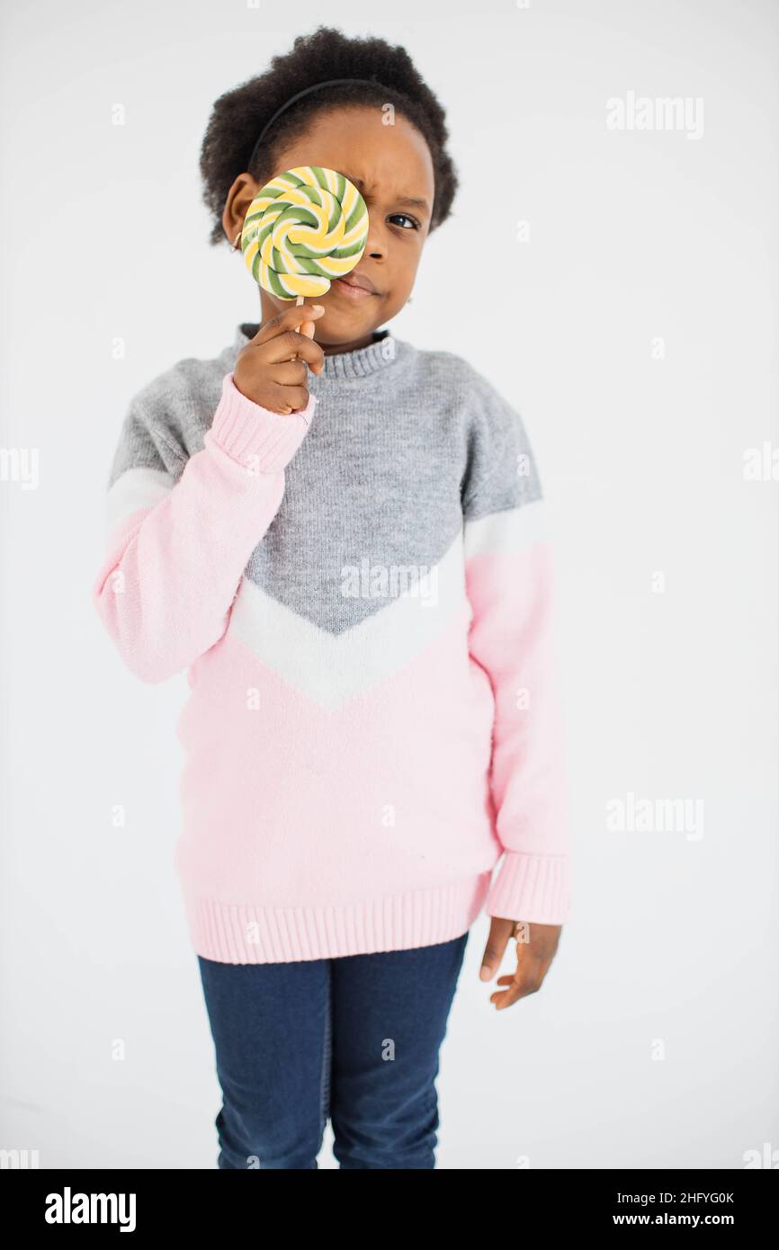 African little child using sweet lollipop for hiding one eye in studio ...