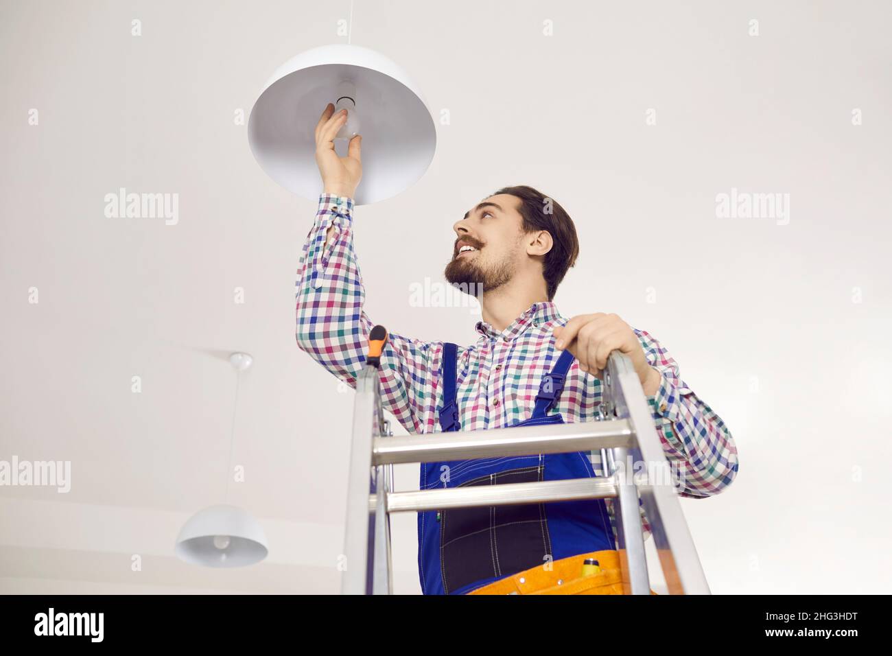 Happy electrician standing on ladder and screwing LED light bulb into ...
