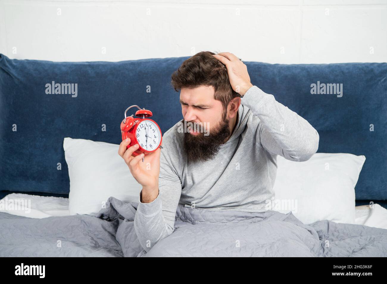 Perplexed guy scratching head holding alarm clock being in bed in ...