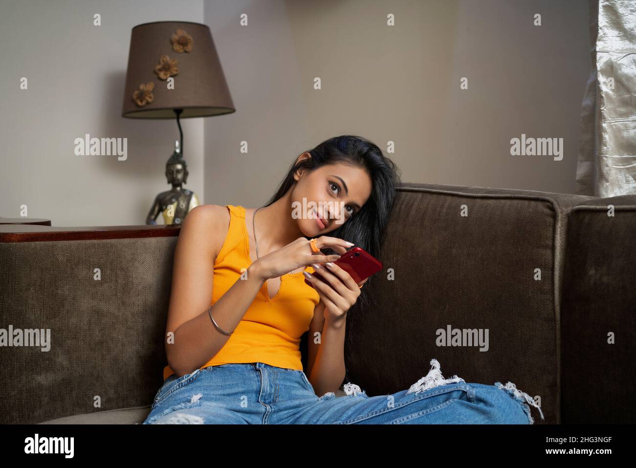 A young woman sitting alone in her room using her mobile phone thinking ...