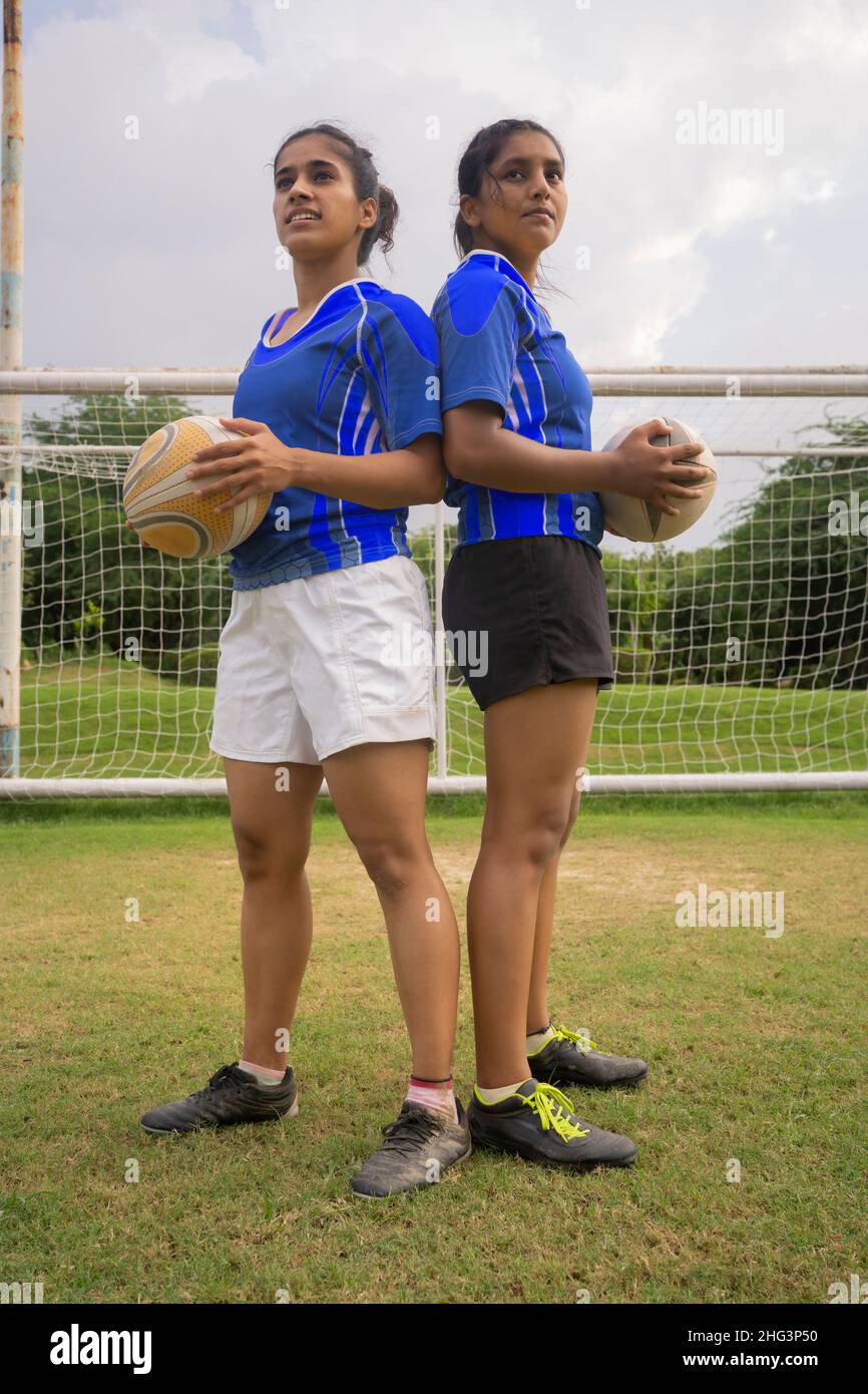 Two young athlete girls standing back to back with rugby balls in front ...