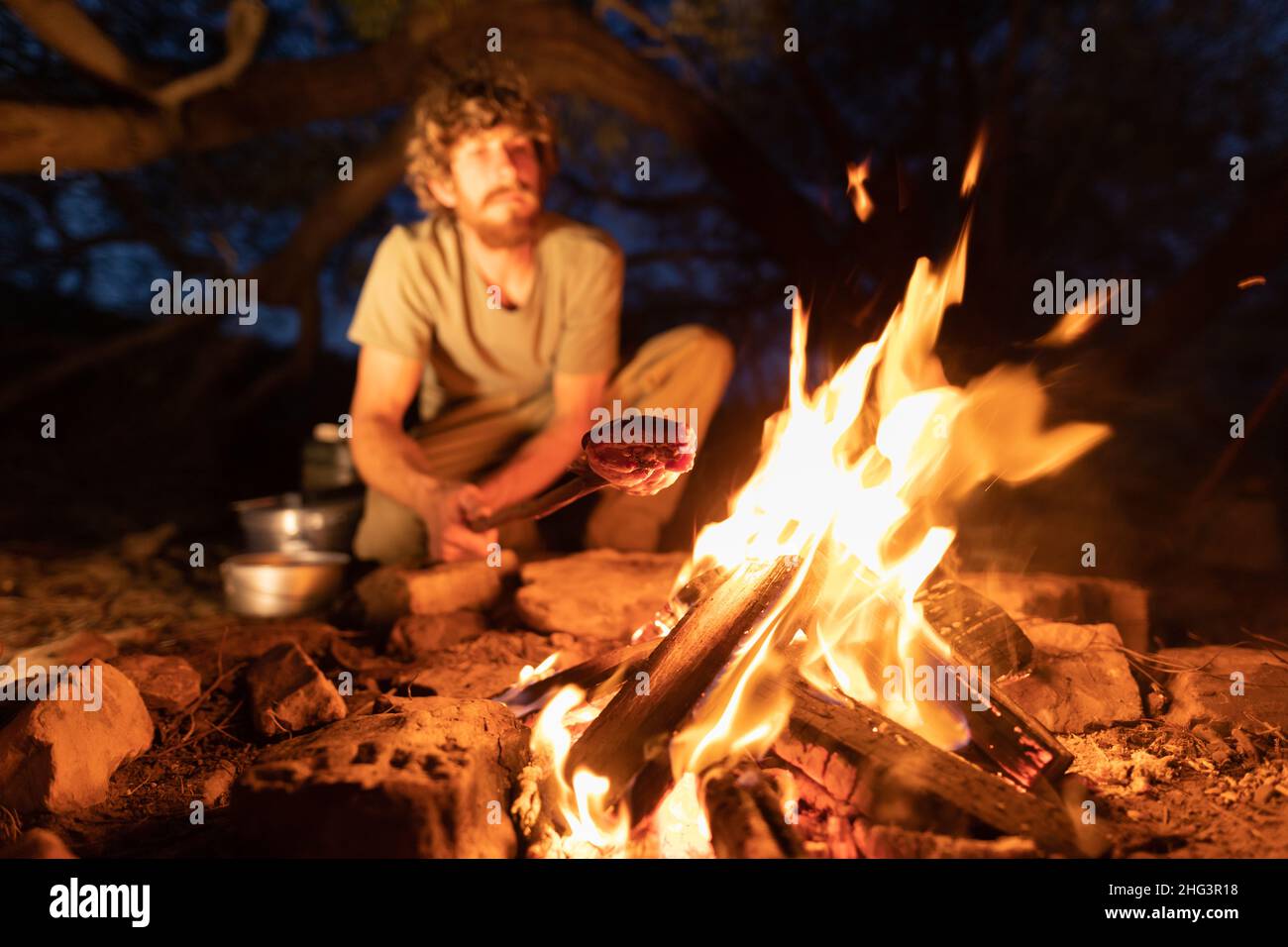 Young male caucasian survivalist cooking meat on burning campfire in ...