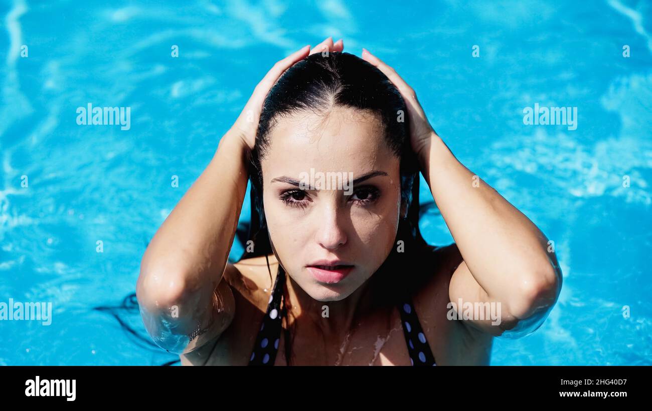 wet and brunette woman adjusting hair while swimming in pool with blue ...