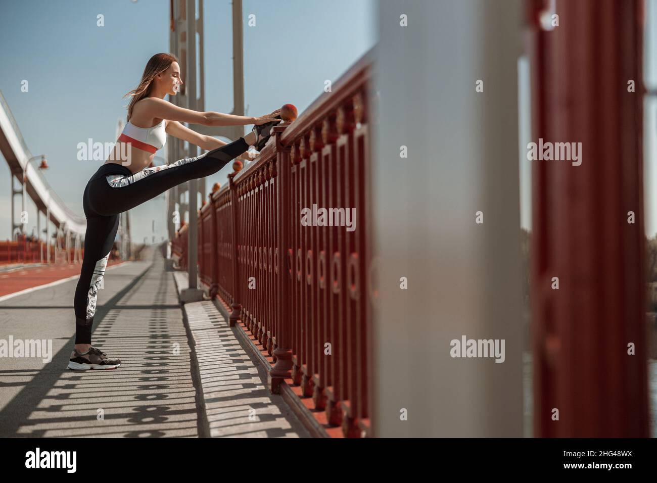 Sportswoman in tracksuit stretches legs leaning onto railing on modern ...
