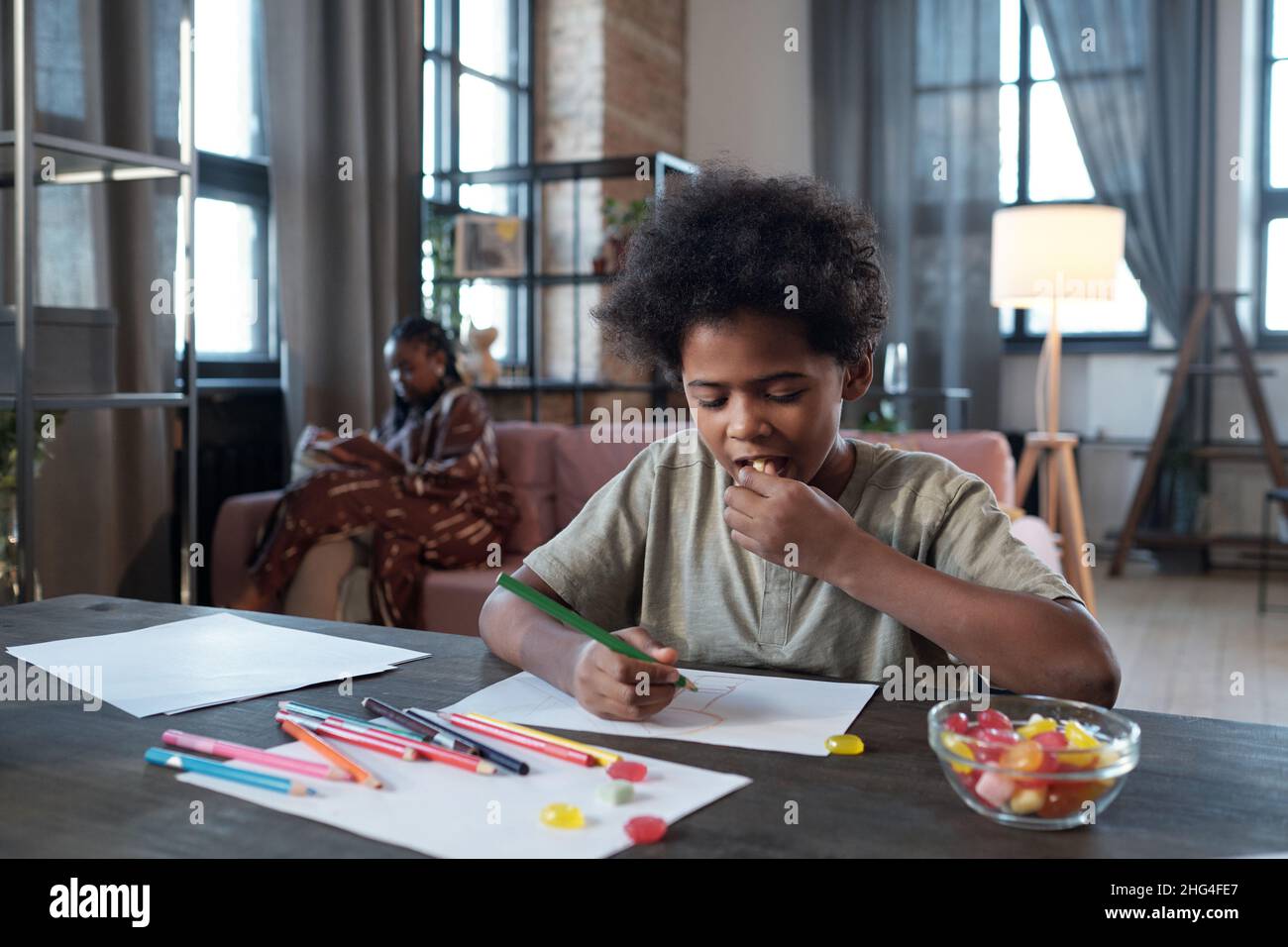 Cute boy eating candies and drawing picture with crayons while sitting ...