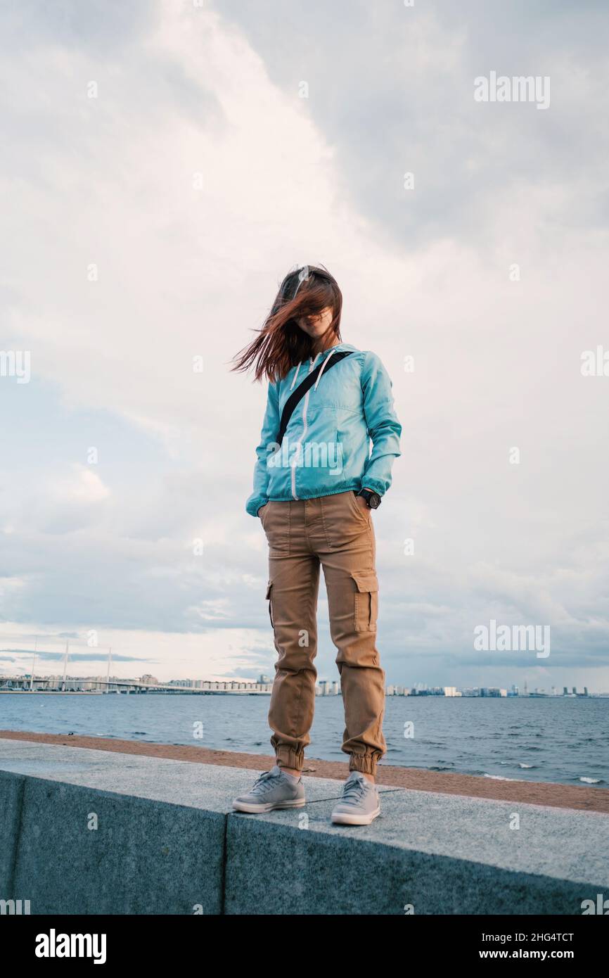 A full-length portrait of a young woman in a blue windbreaker in windy ...