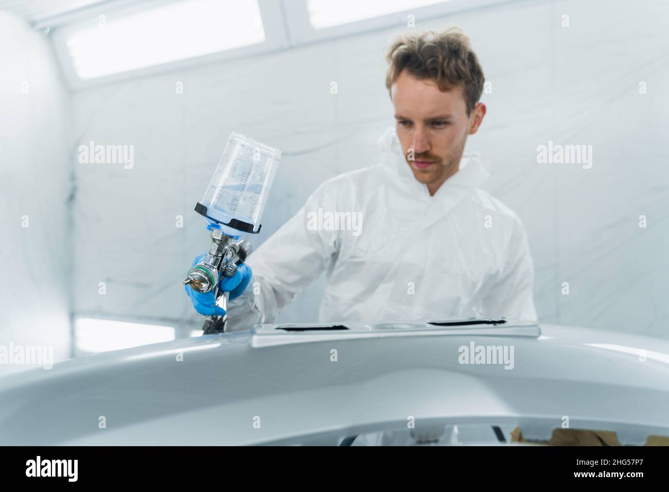 Young curly car painter varnishes a car bumper. Man works in a spray ...