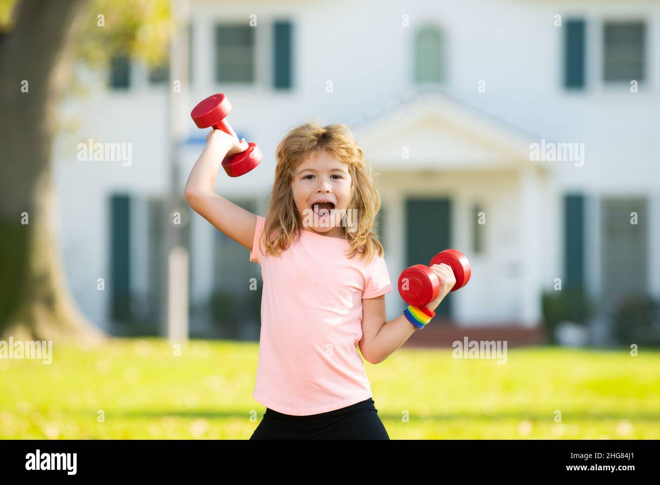 Cute excited child boy pumping up arm muscles with dumbbell outdoor in summer park. Fitness kids ...