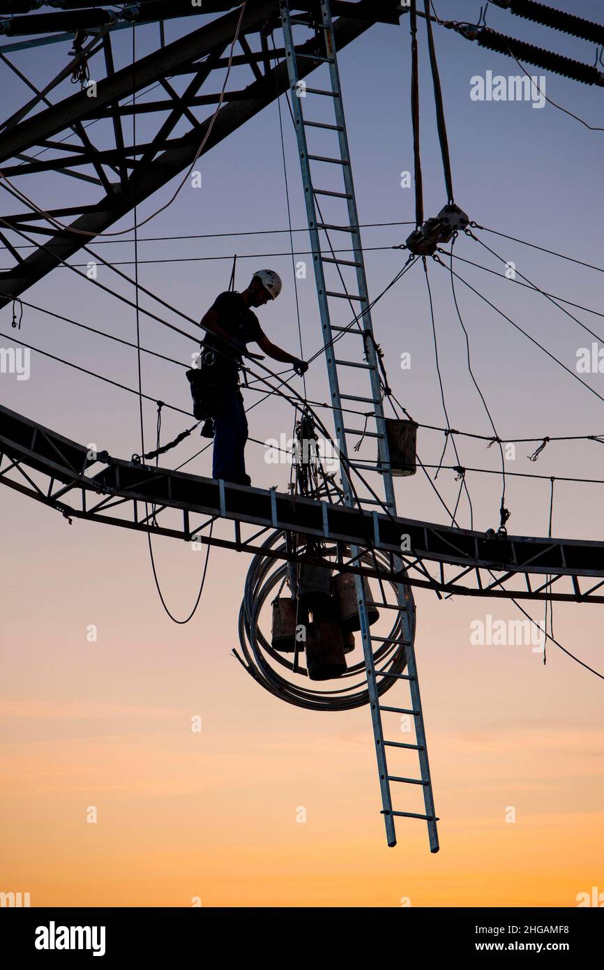 High-voltage fitter at work, backlit by evening sky, overhead power ...
