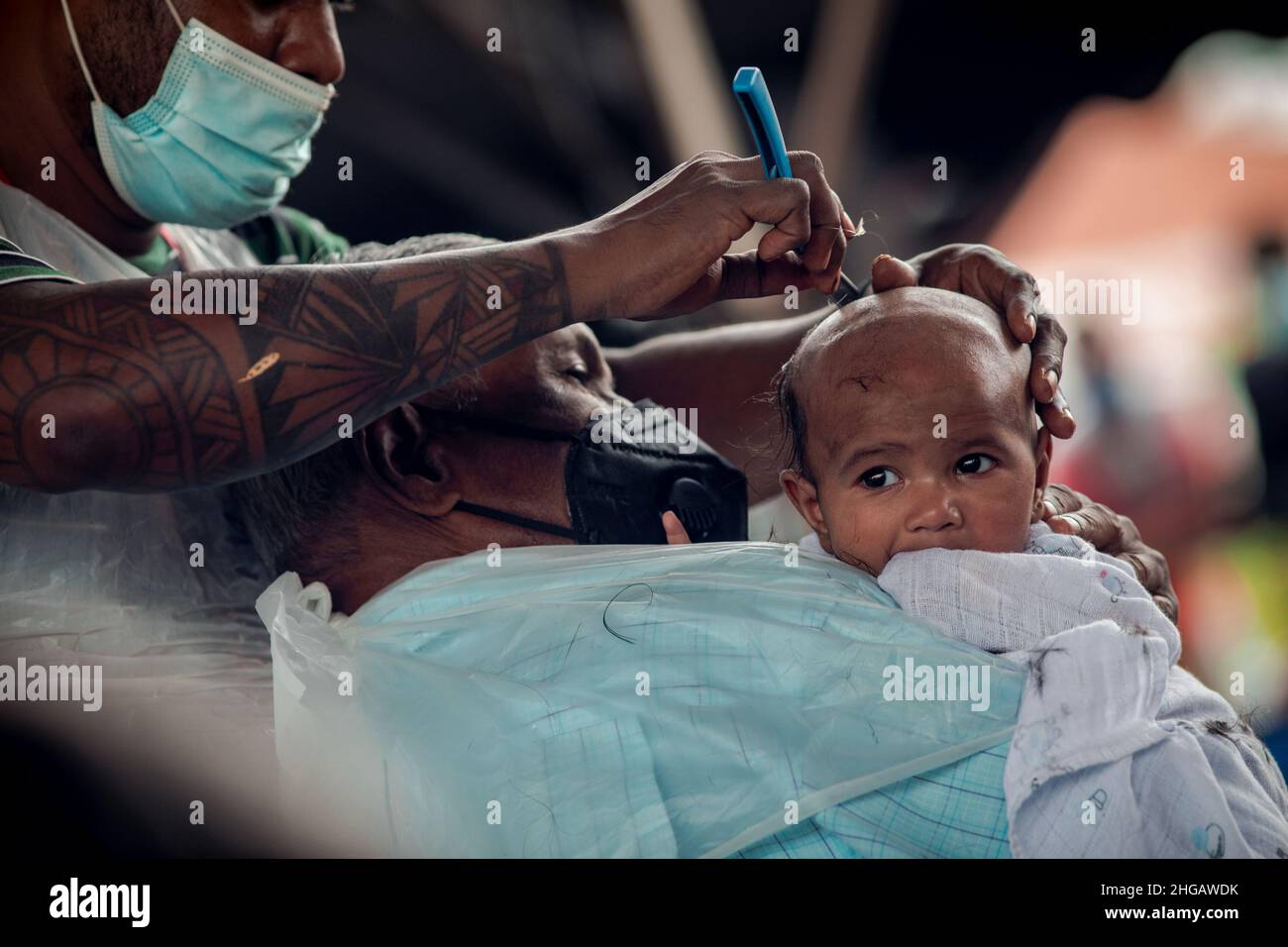 A barber wearing a face mask is seen shaving a baby's head to complete ...