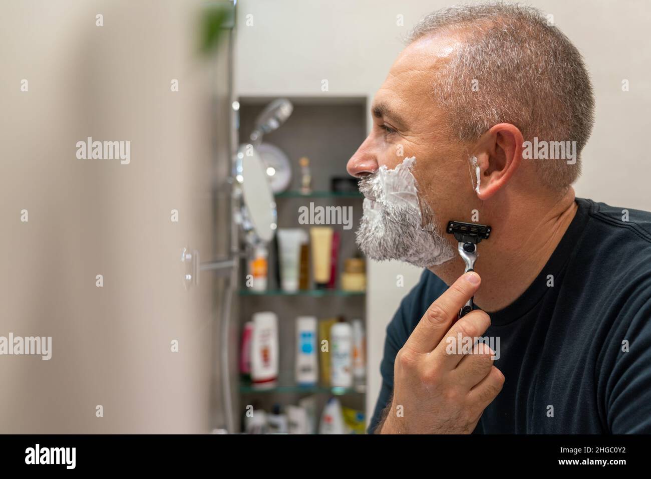 Handsome white-haired beared man shaving off his beard looking in a ...