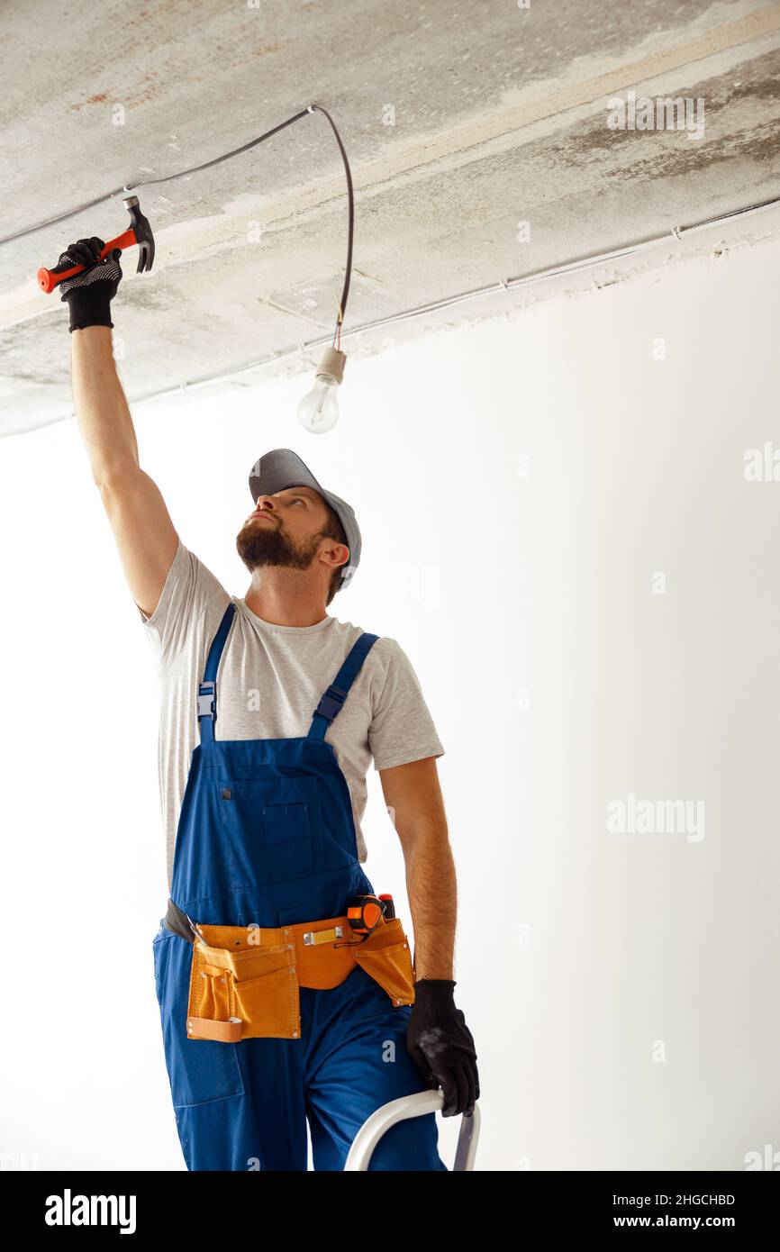 Focused electrician in uniform using hammer while attaching wire to ...