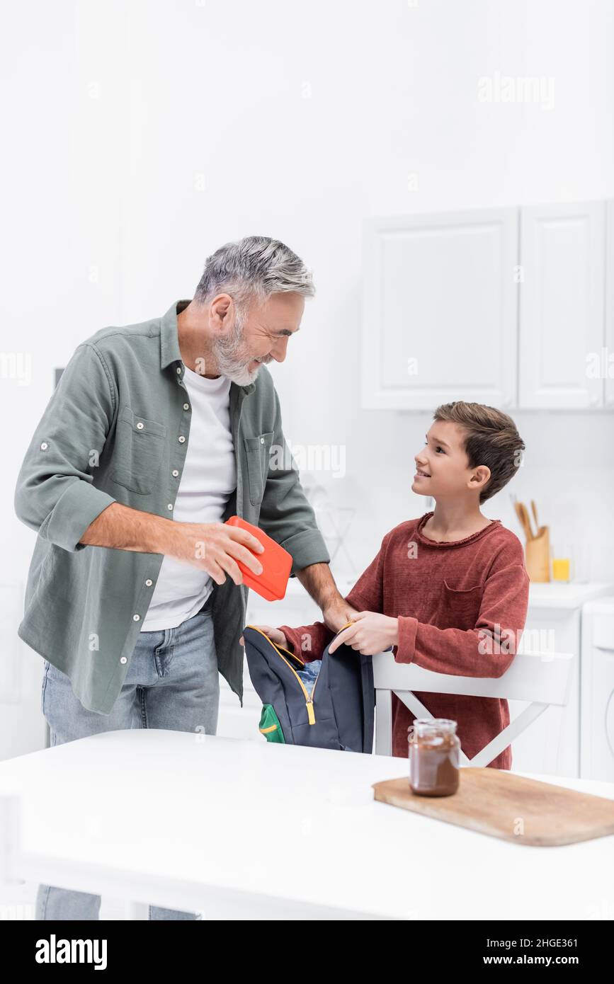 grey-haired man holding lunch box while packing backpack of smiling ...