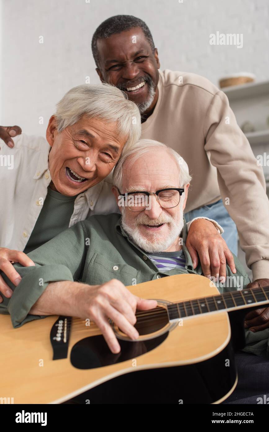 Cheerful interracial senior friends playing acoustic guitar and hugging ...