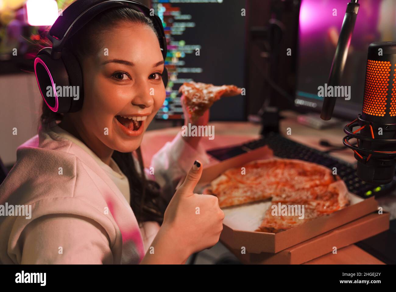 Smiling young woman gmaer in headphones eating pizza while sitting at ...