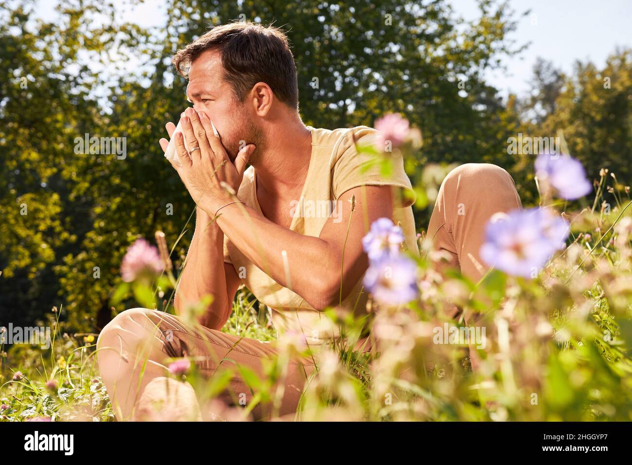 Man with pollen allergy sits among blooming flowers and blows his nose ...