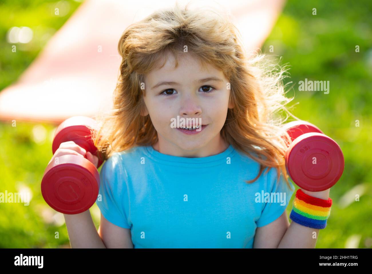 Cute child boy pumping up arm muscles with dumbbell outdoor in summer park. Fitness kids with ...
