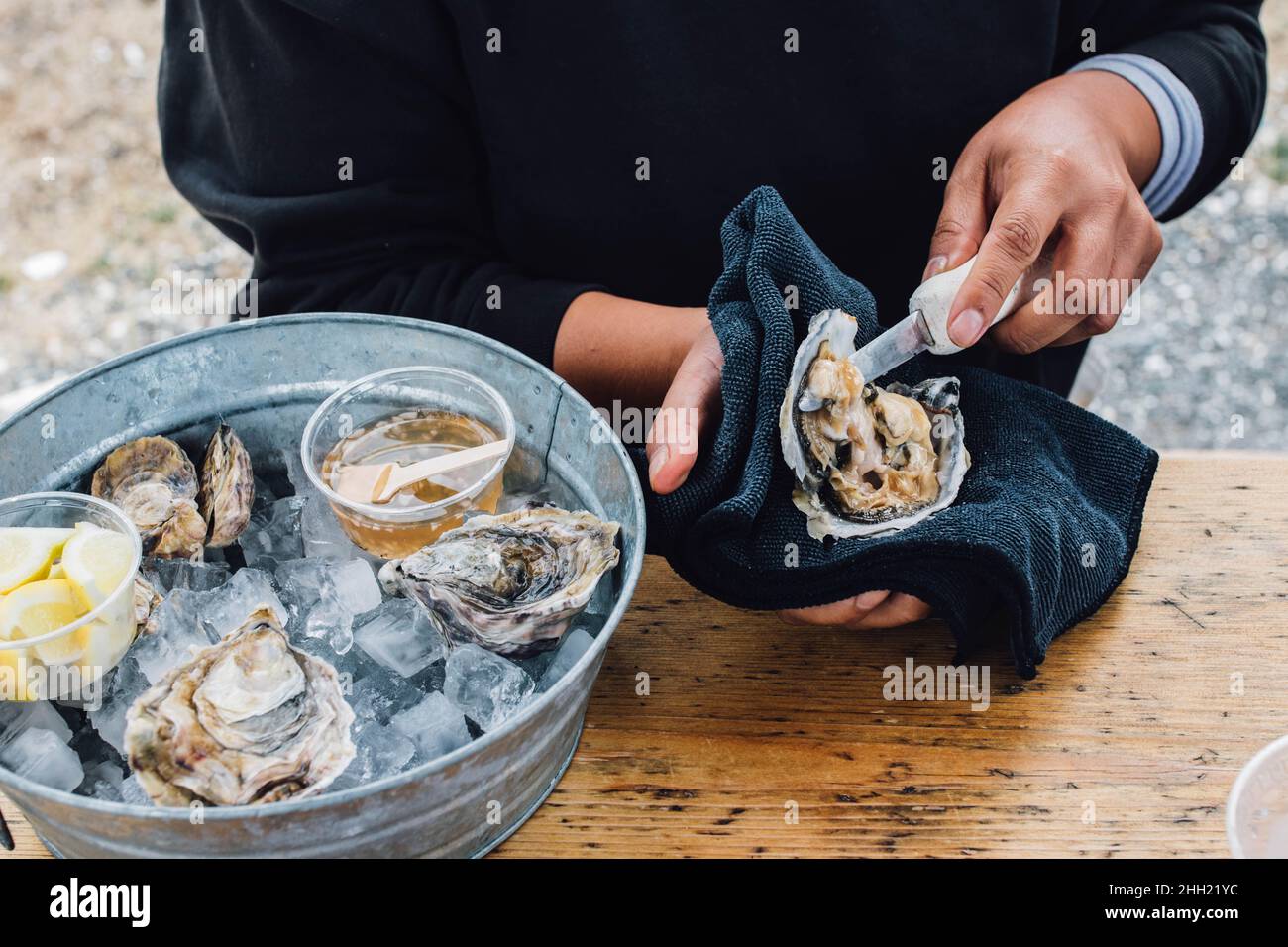 woman shucking oyster at table outdoors with ice-filled tub of oysters ...