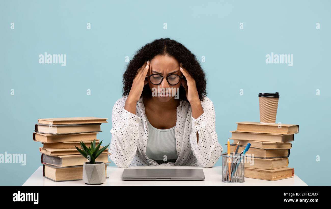 Exhausted black female student studying at table with laptop and heaps ...