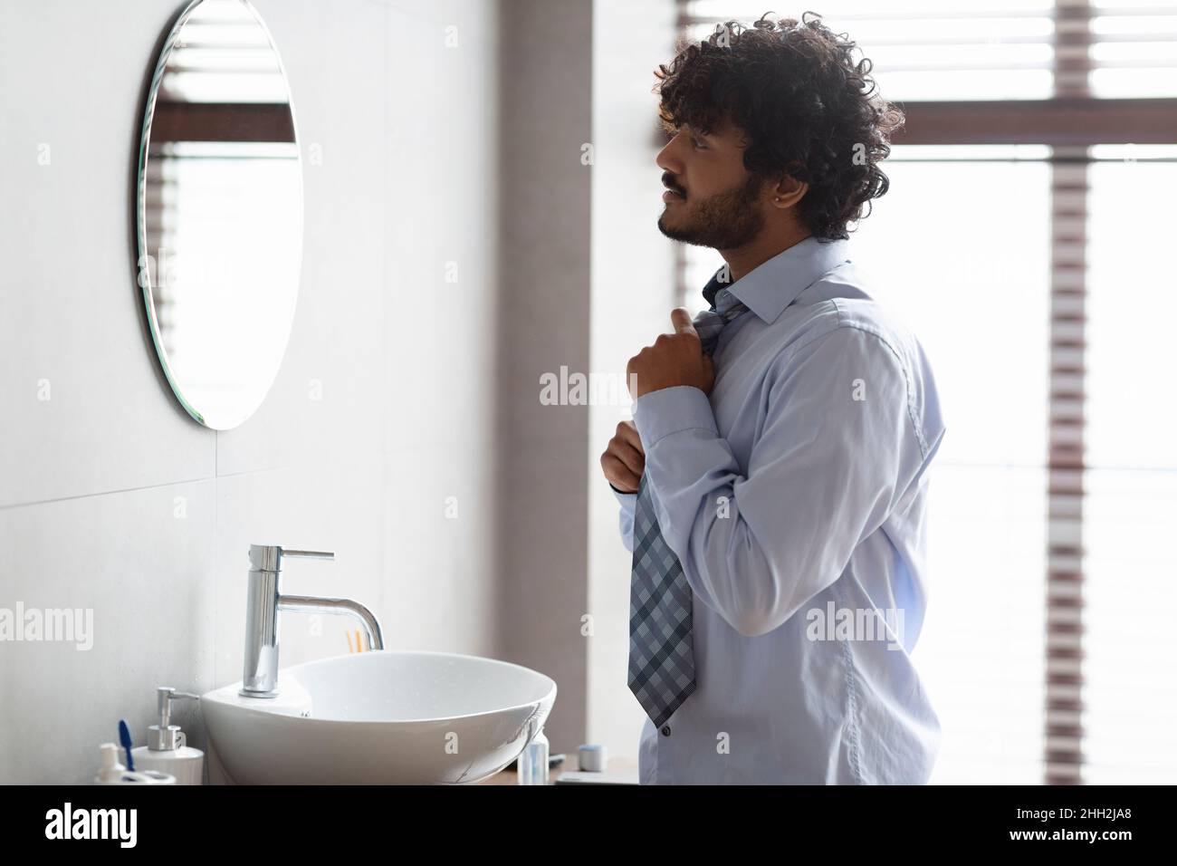 Young indian businessman tying necktie and preparing for work, standing ...