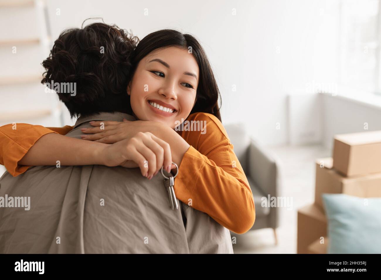 Smiling young Asian woman hugging her boyfriend and holding house key on moving day, free space ...