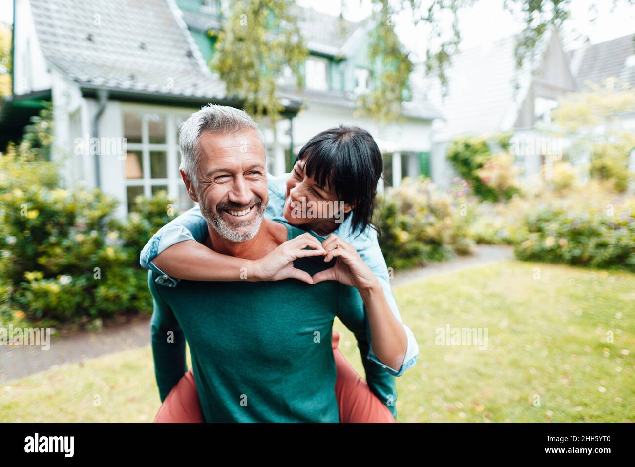 Cheerful man giving piggyback ride to woman gesturing heart shape in ...
