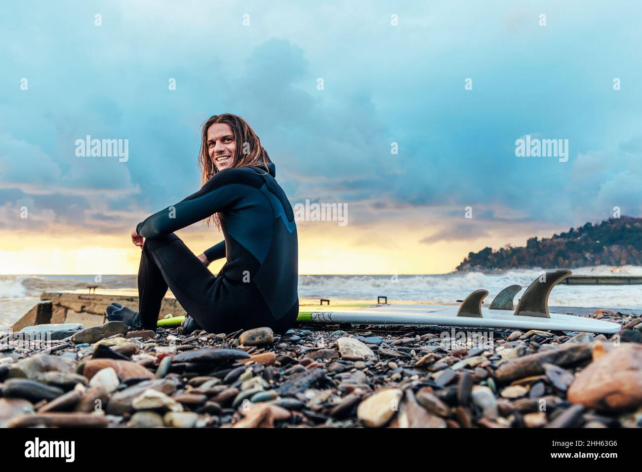 Smiling surfer looking over shoulder sitting by surfboard at beach ...