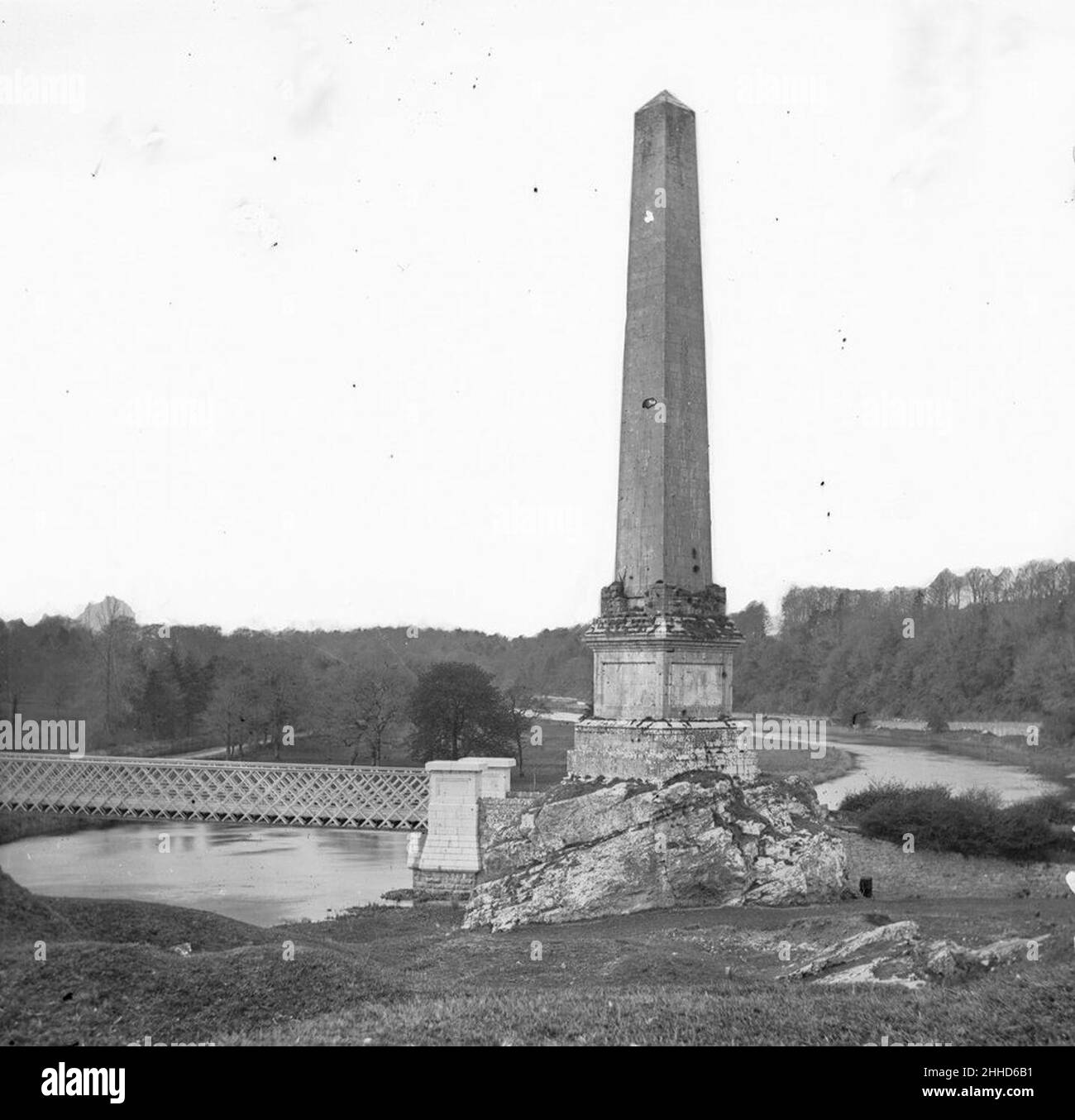Square obelisk by footbridge with iron railing over wide stream, woods ...