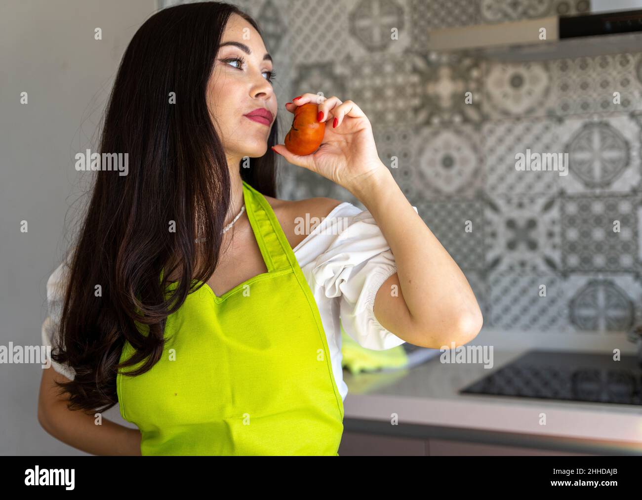 Trendy woman in green apron looking away and smelling ripe tomato while ...