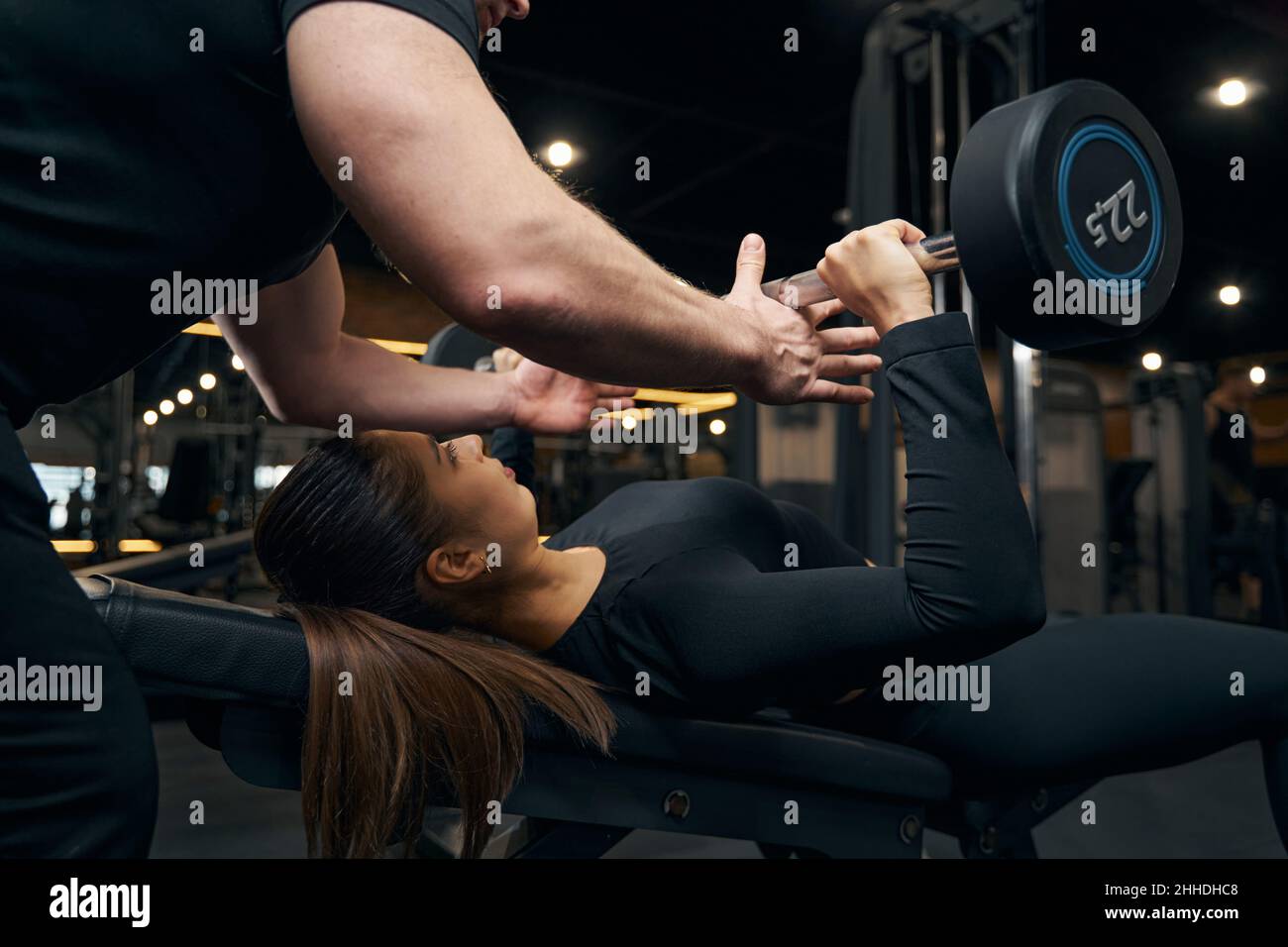 Female lifting barbell with both hands supported by trainer Stock Photo ...