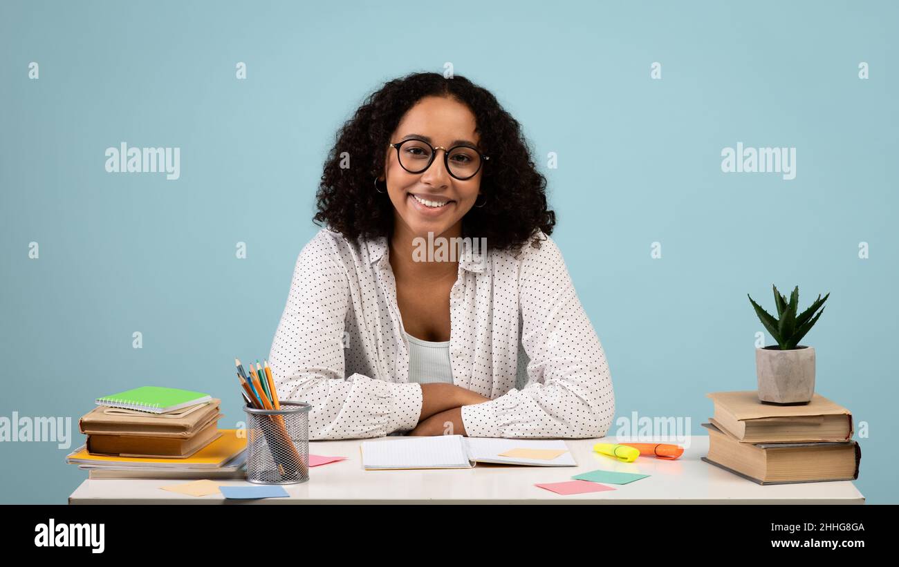 Happy young black female student sitting at desk with books and ...
