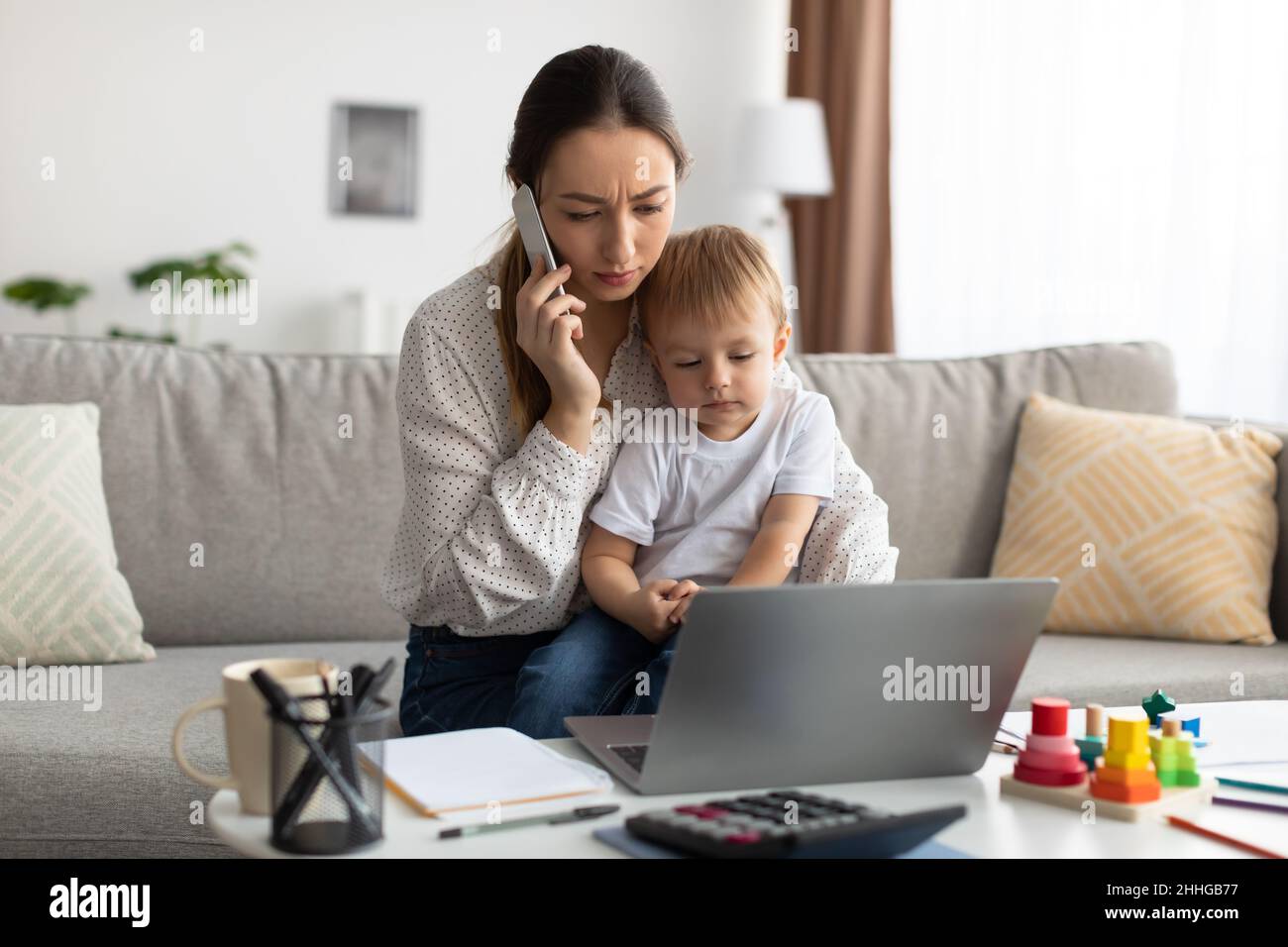 Millennial mother working on laptop computer and talking on phone ...