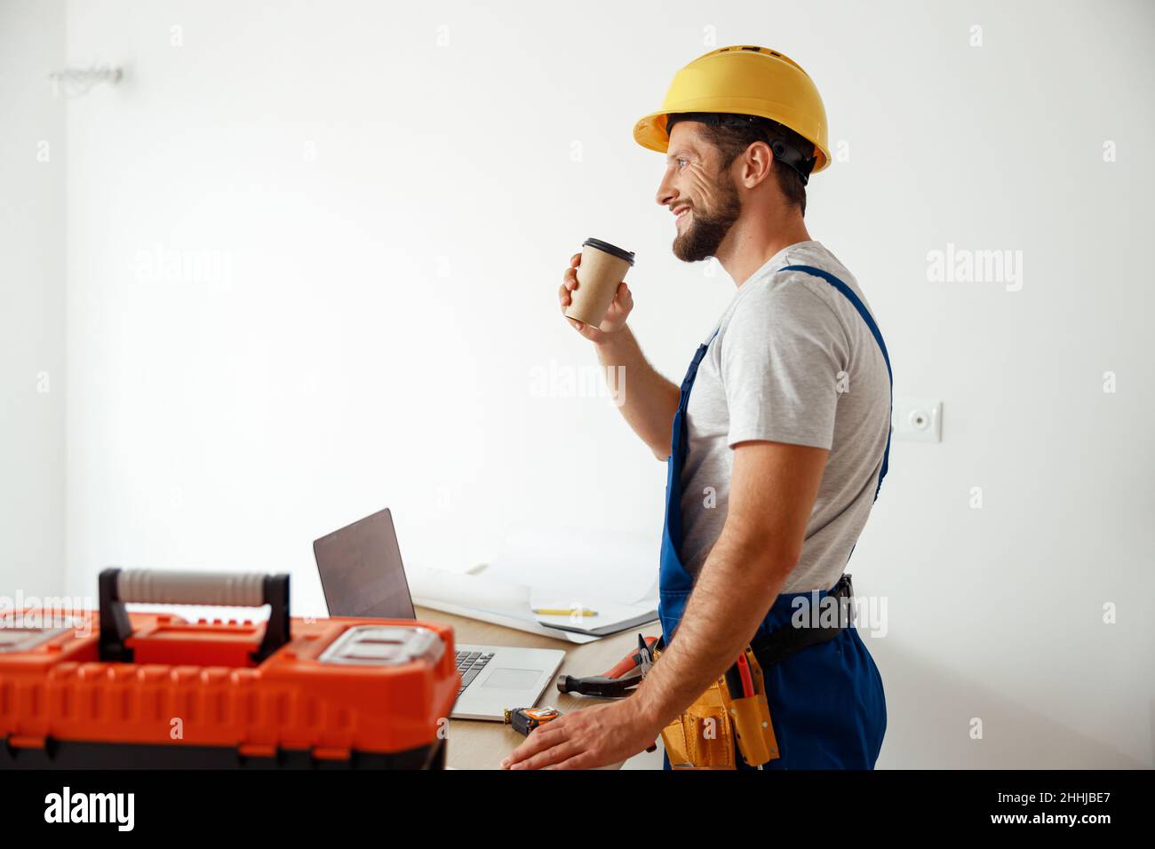 Side view of smiling handyman in uniform and hard hat drinking coffee ...