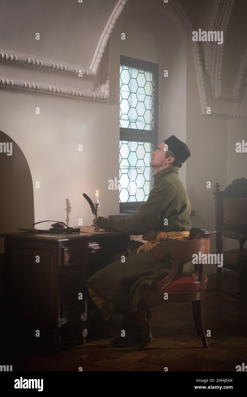 Portrait of a man in a medieval costume. A scribe writes on a red table ...