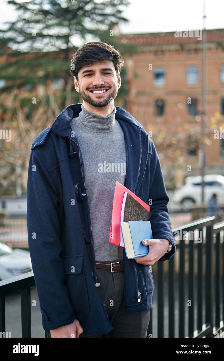 Positive male student carrying books for studies on walkway in urban ...