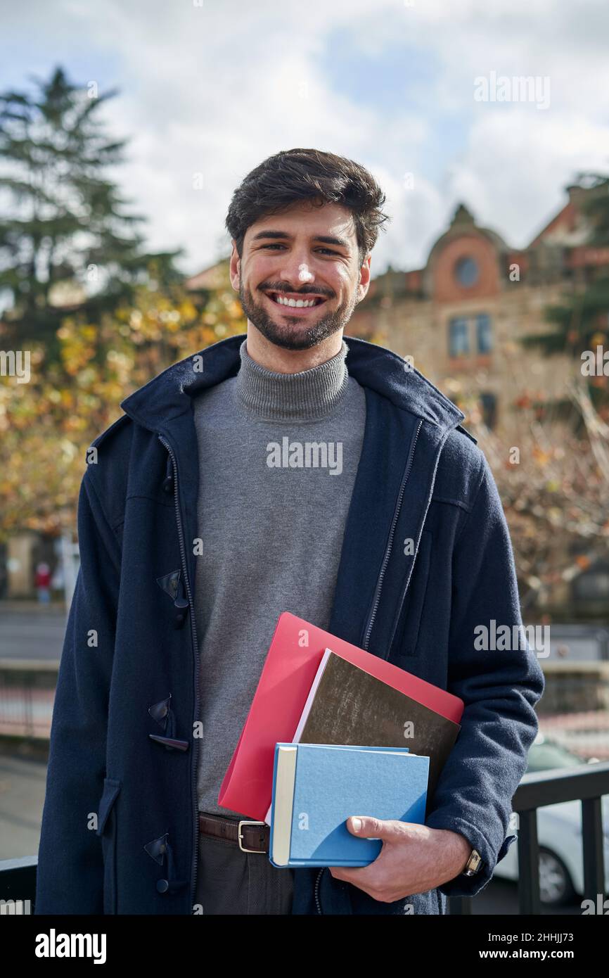 Positive male student carrying books for studies on walkway in urban ...