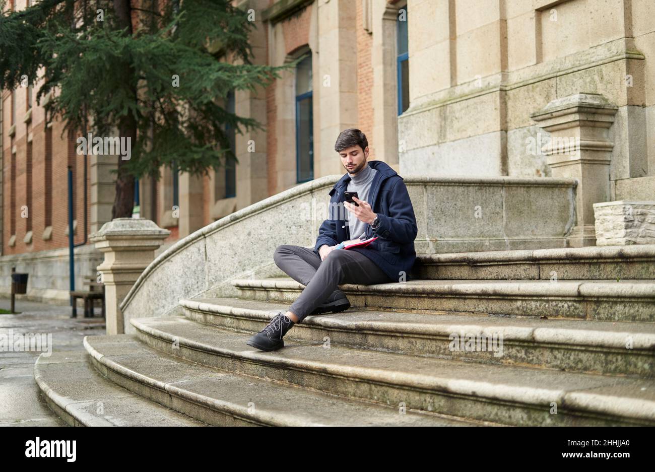 Full body of serious bearded male student with notepad sitting on stone ...