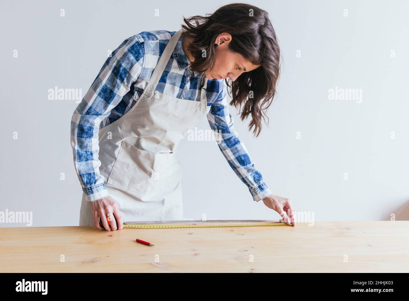 Concentrated female artisan in apron measuring wooden plank with ...
