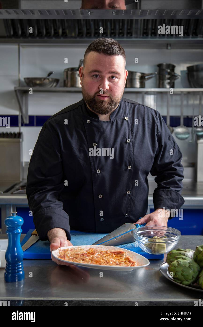 Bearded male chef in black uniform looking at camera and offering plate ...