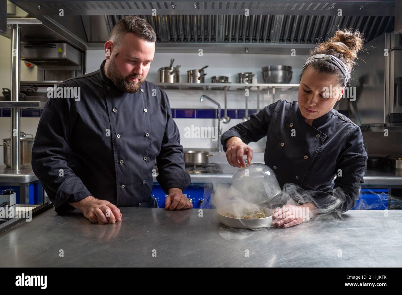 Male and female chefs in dark uniform removing dome lid from tasty dish ...