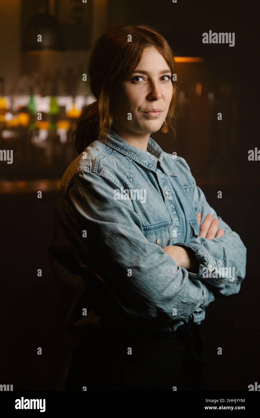 Attractive waitress in apron looking at camera with crossed arms while ...