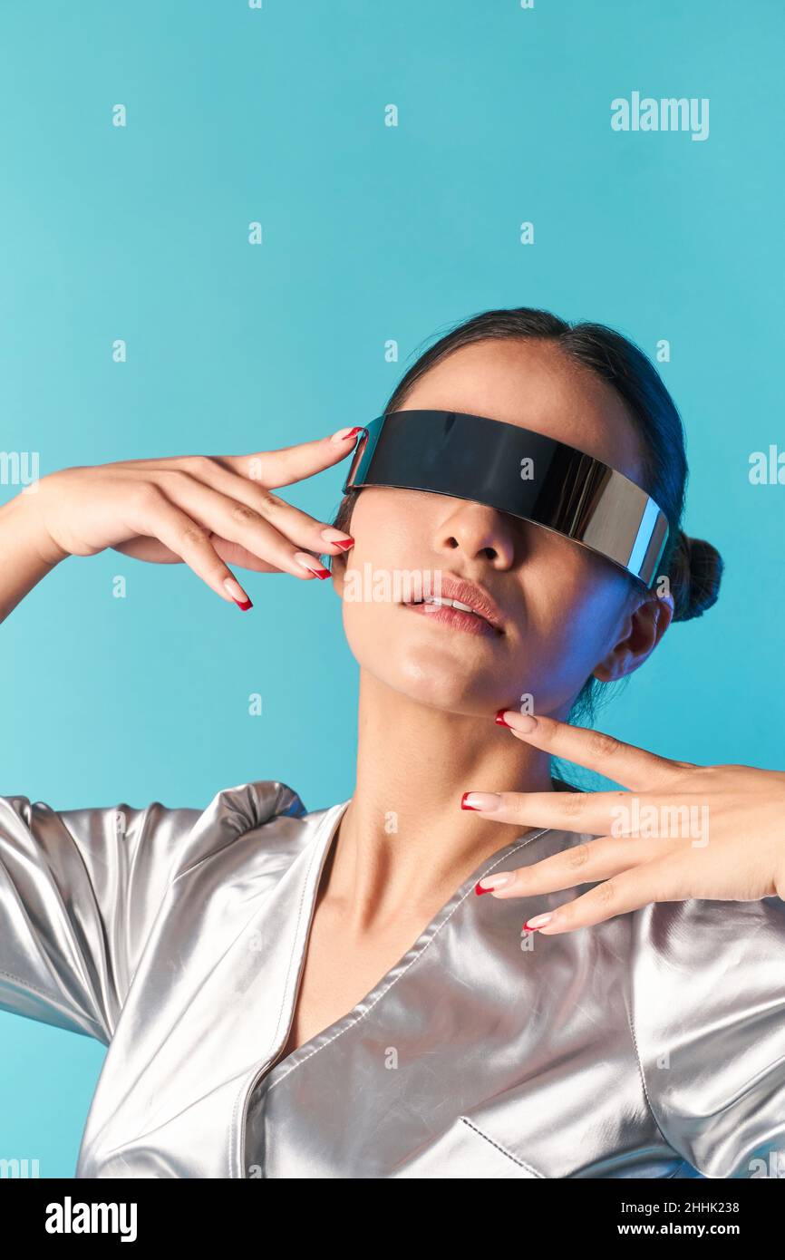Young trendy ethnic lady with dark hair in modern mirrored VR goggles touching face against blue ...