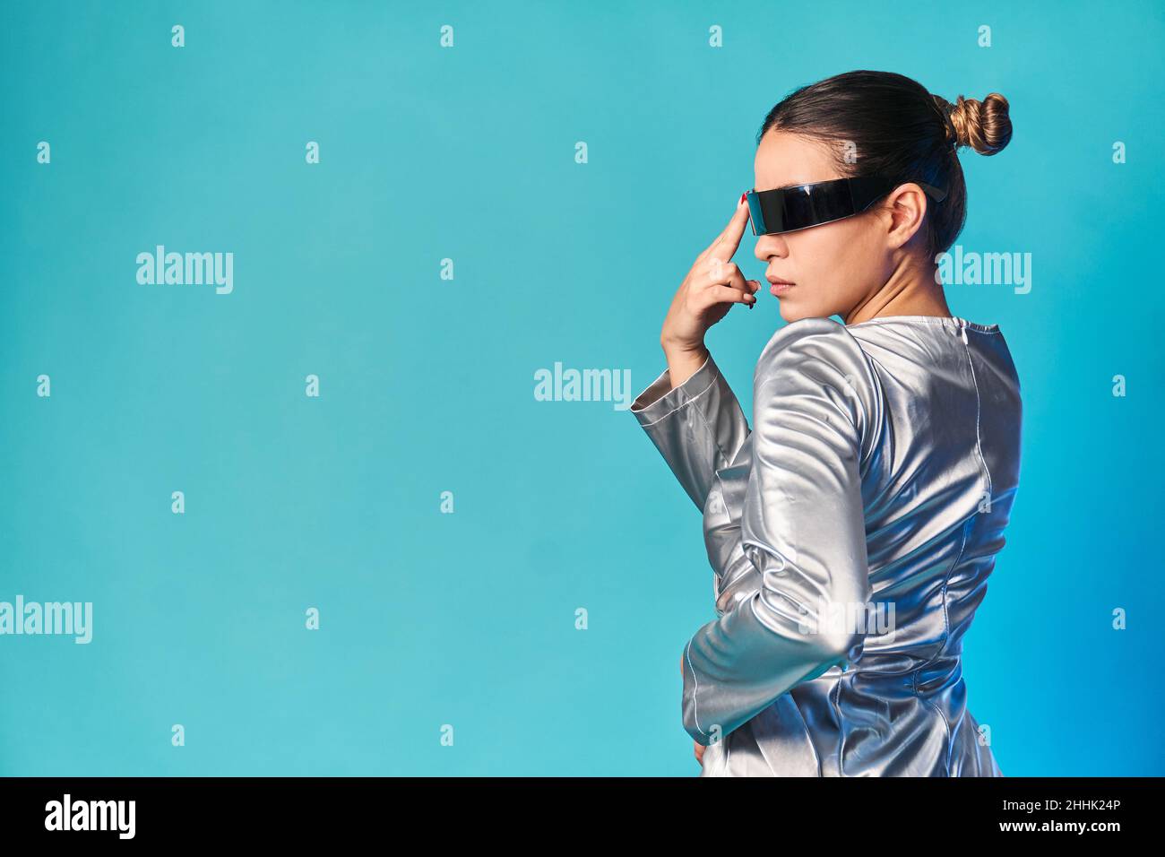 Young trendy ethnic lady with dark hair in modern mirrored VR goggles touching face against blue ...